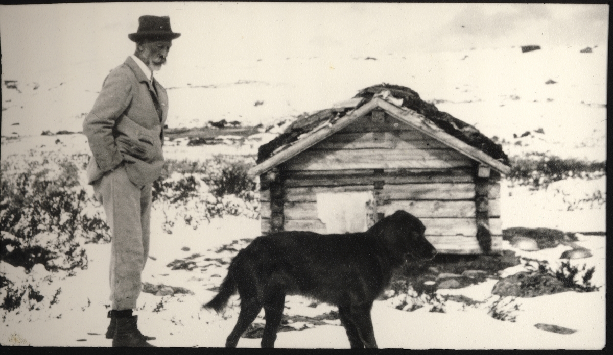 Ferdinand Egeberg og hund foran et lite, laftet uthus. Snø dekker ...