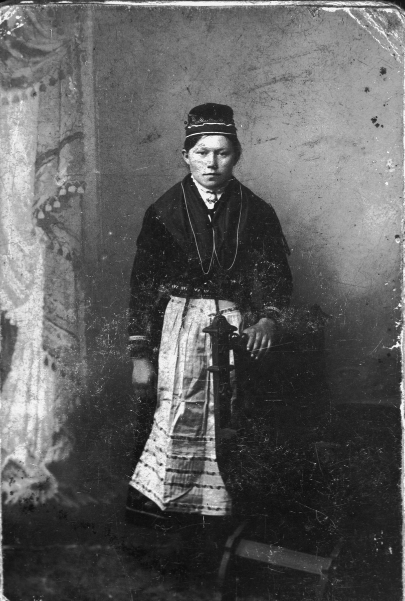 Studioportrett av ung kvinne i samisk drakt stående ved en stol. - Sør ...