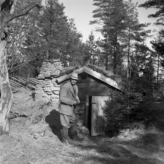 En backstuga med tak av mossa. Framför står en man klädd i hatt och ...